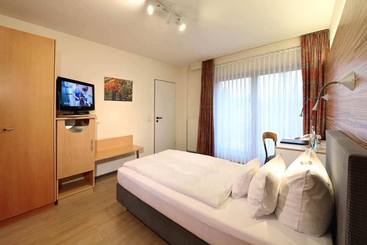 Quarto de hotel com uma cama, televisão, escrivaninha e janela com cortinas.