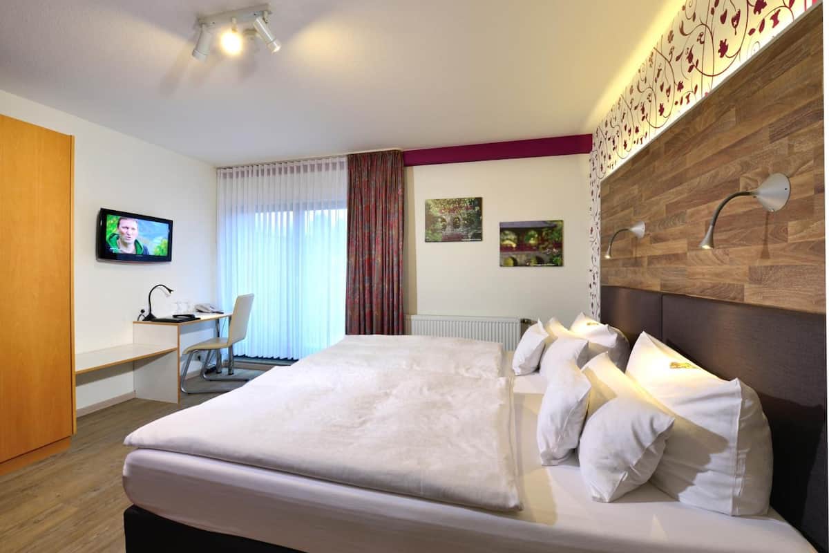 Quarto de hotel com cama, escrivaninha com cadeira, televisão e janela com cortinas.