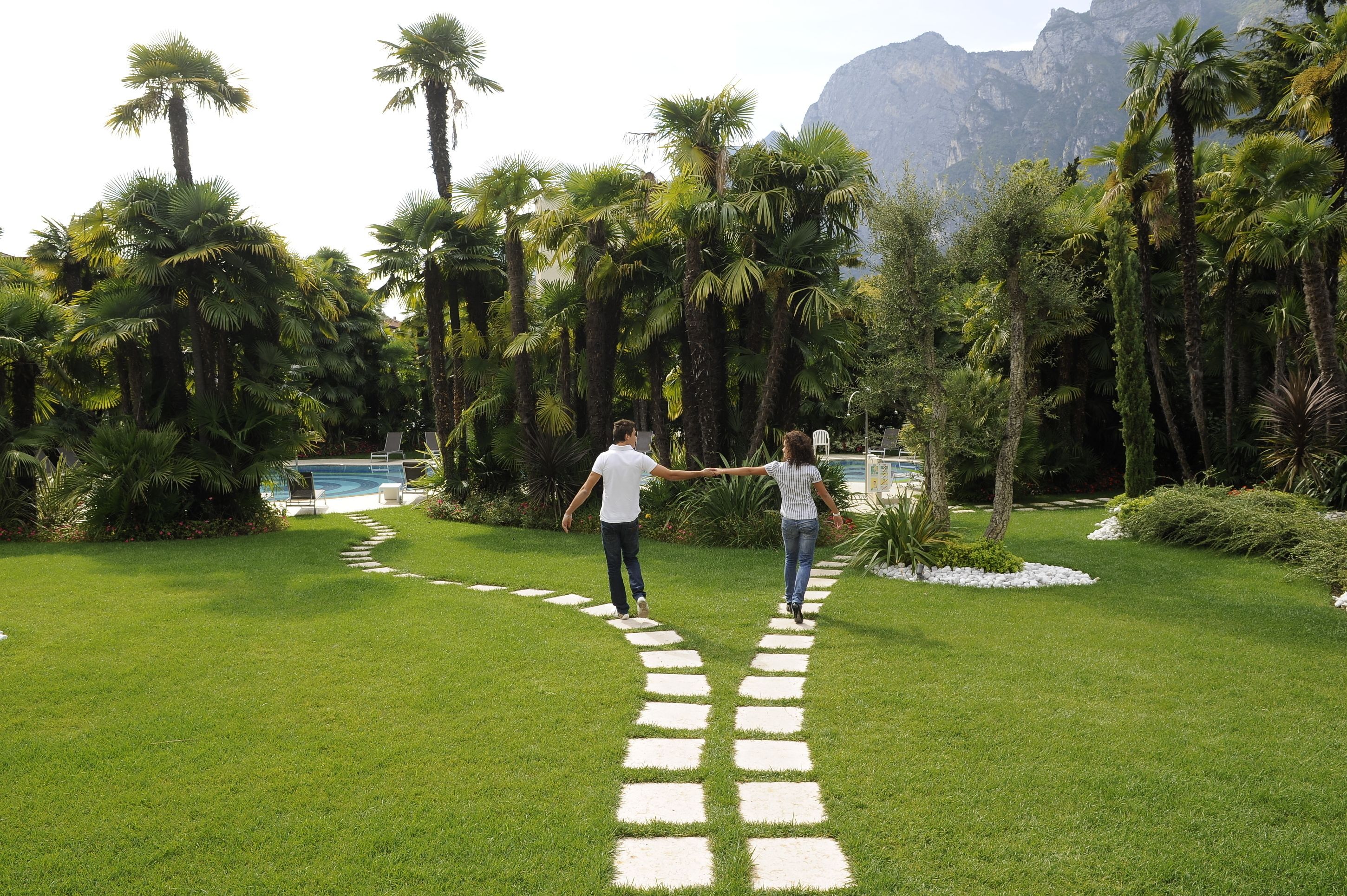 Um casal caminhando por um caminho de pedra em um jardim exuberante, com palmeiras e uma montanha ao fundo.