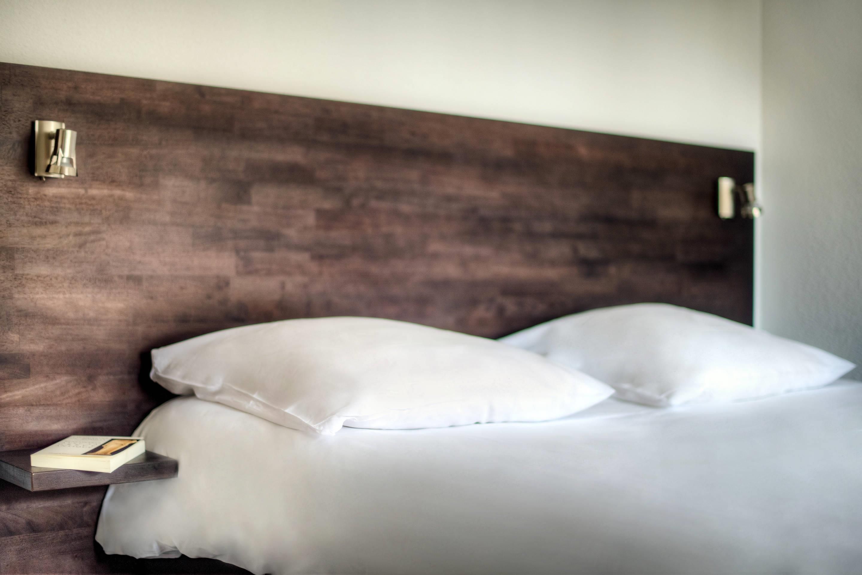 Cama com roupa de cama e almofadas brancas, cabeceira de madeira e uma pequena mesa de cabeceira com um livro.