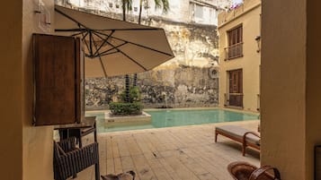 Uma área à beira da piscina com piso de azulejo, espreguiçadeiras, uma cadeira de vime e um grande guarda-sol.
