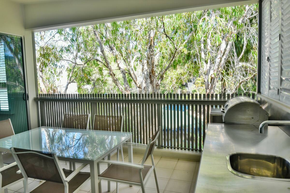 Varanda com mesa e cadeiras de vidro, pia de aço inoxidável e vista para árvores.