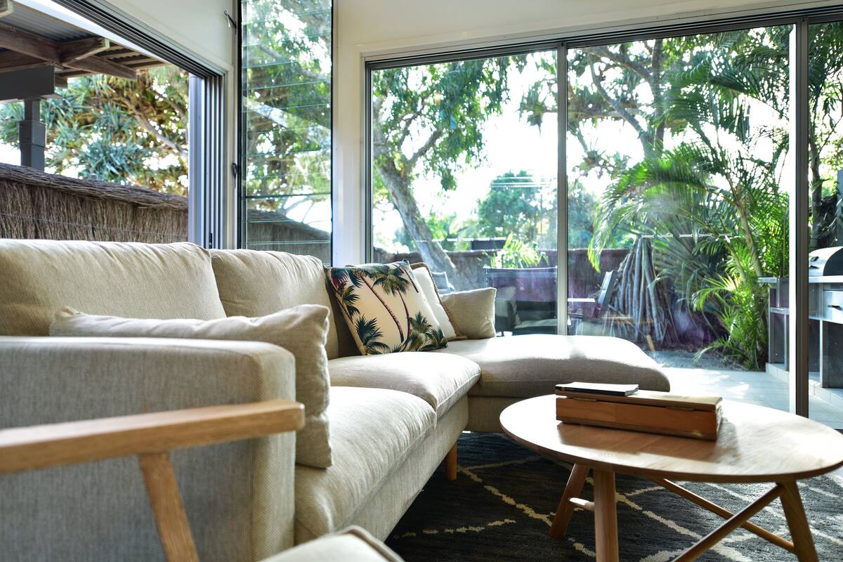 Uma sala de estar moderna com um sofá, uma mesa de centro redonda e vista para um jardim exuberante através de grandes janelas.