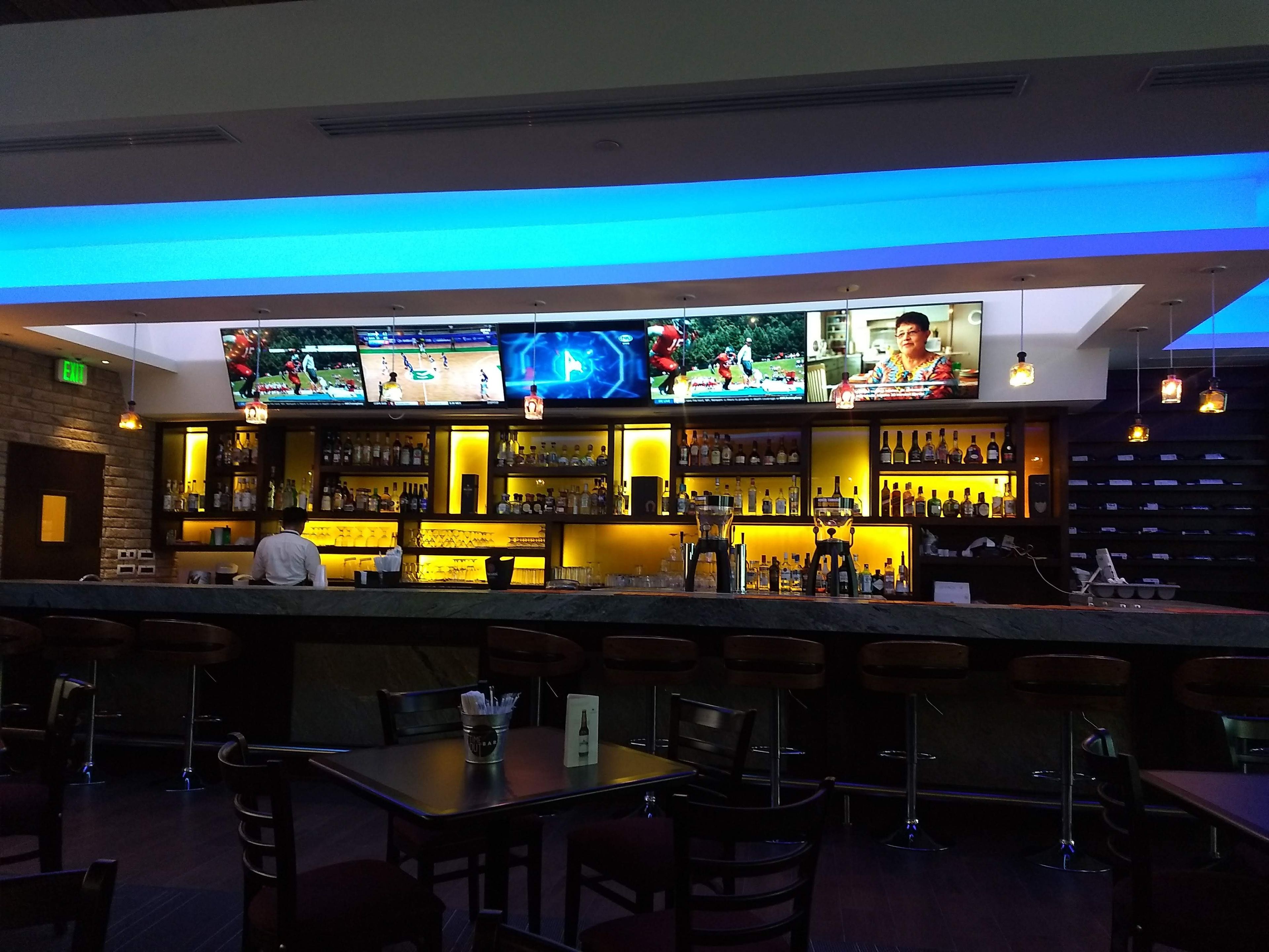 Espaço de bar com várias telas exibindo esportes, um bartender e uma variedade de garrafas nas prateleiras.