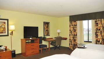 Quarto de hotel com cama, escrivaninha, cadeira, televisão e janela com cortinas.