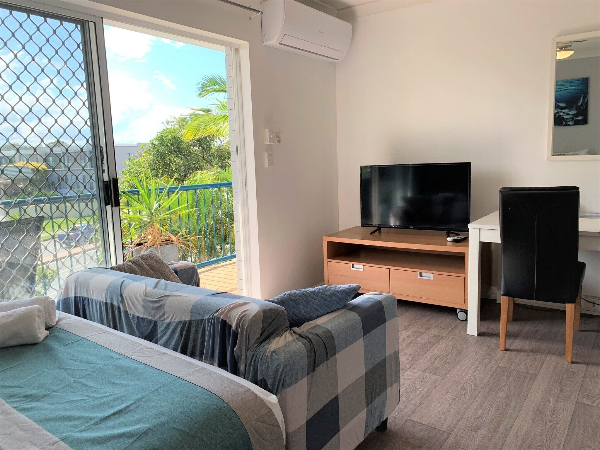 Quarto com cama, escrivaninha, cadeira, televisão e varanda com vista para vegetação.