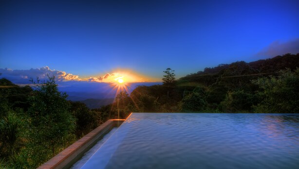 Piscina com vista para o pôr do sol sobre uma paisagem montanhosa.