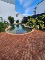 Piscina retangular com borda de tijolos, rodeada por vegetação e paredes brancas.