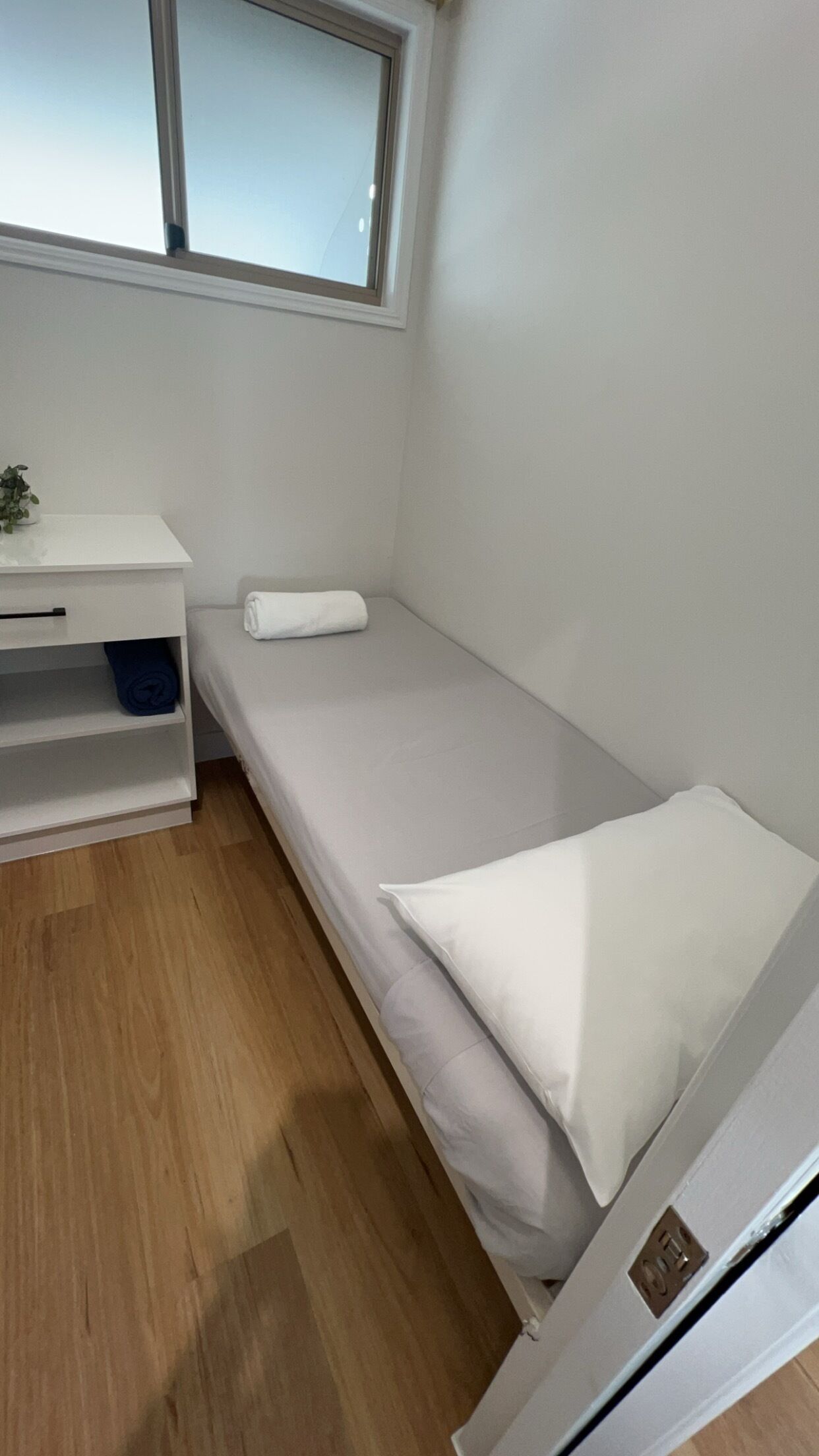 Quarto pequeno com uma cama de solteiro, uma escrivaninha com prateleira e uma janela.