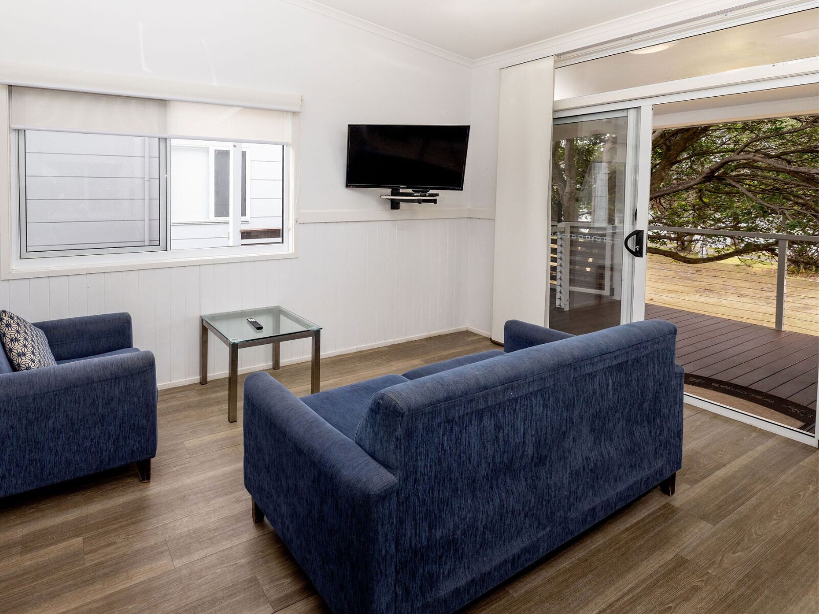 Uma sala de estar moderna com uma televisão grande, dois sofás, uma mesinha e uma porta de vidro deslizante que dá para uma área externa.