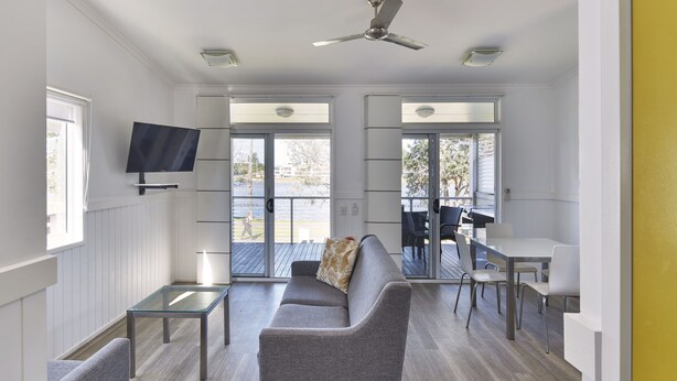 Uma sala de estar moderna com um sofá cinza, uma mesa de centro de vidro e uma TV de tela plana na parede. Há um ventilador de teto, um espaço de jantar com mesa e cadeiras, e grandes janelas que oferecem vista para o exterior.