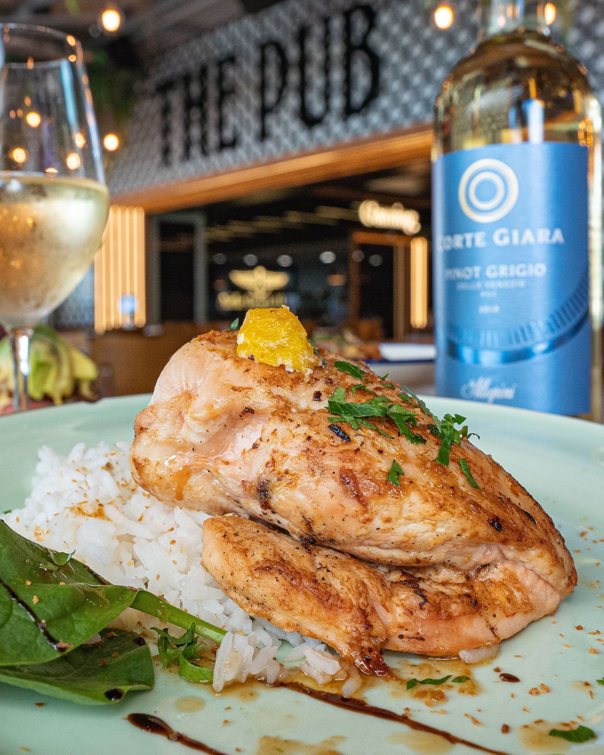 Prato de frango grelhado com arroz e guarnição, uma garrafa de vinho e uma placa com a inscrição 'THE PUB' ao fundo.