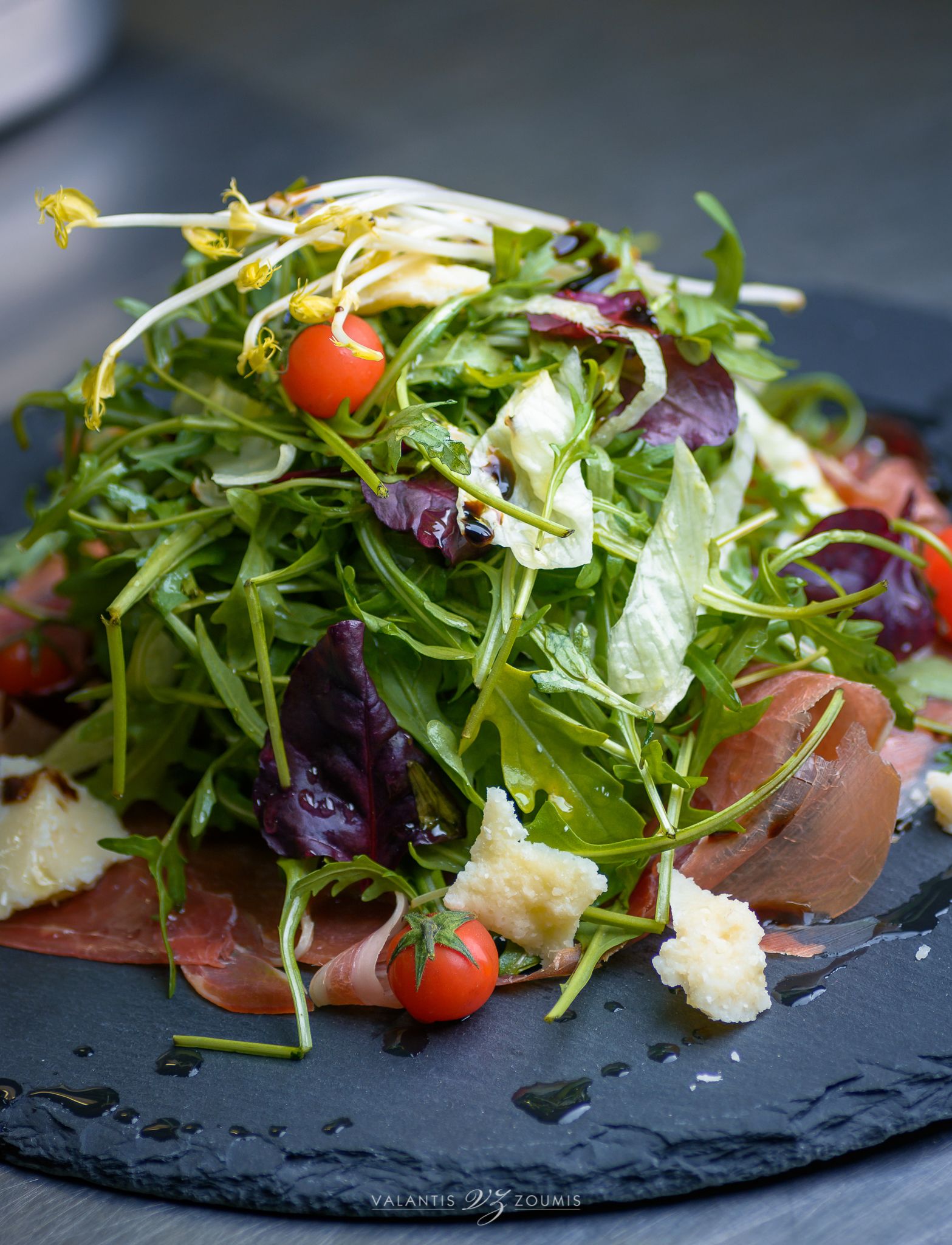 Salada com diversas folhas, tomates cereja e queijo sobre um prato de ardósia.