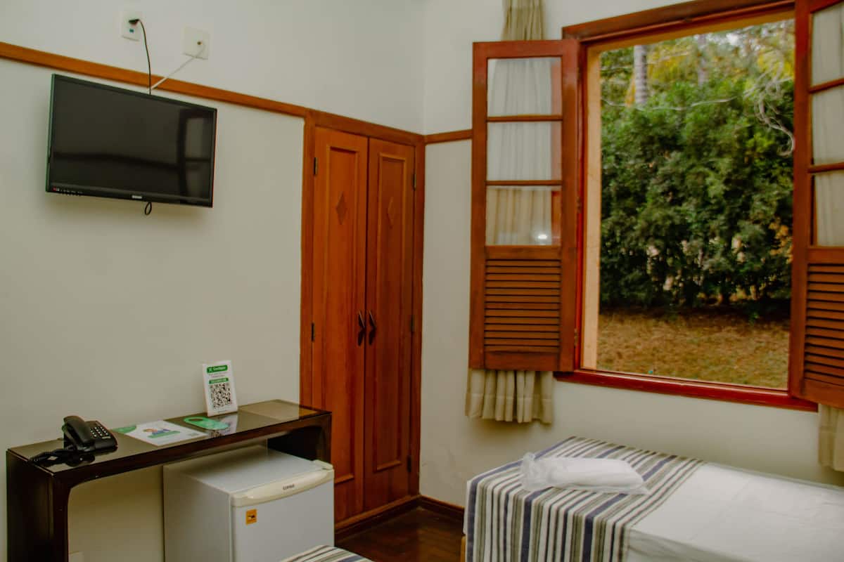 Quarto com cama, escrivaninha com computador, telefone e janela com vista para o verde.