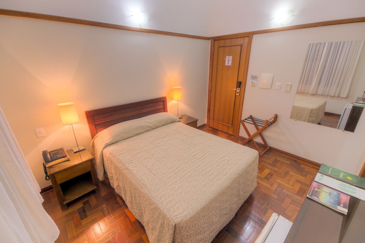 Quarto bem arrumado com cama, luminárias de cabeceira, mesa de cabeceira de madeira, uma cadeira, uma porta e uma pequena mesa com livros.
