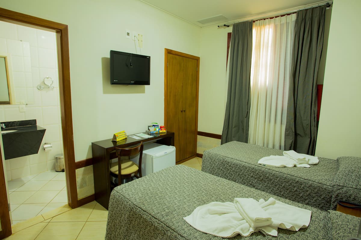 Quarto de hotel com duas camas, uma escrivaninha com cadeira, uma televisão e um banheiro visível através de uma porta aberta.