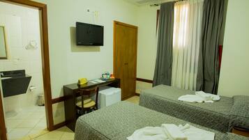 Quarto de hotel com duas camas, uma escrivaninha com cadeira, uma televisão e um banheiro visível através de uma porta aberta.