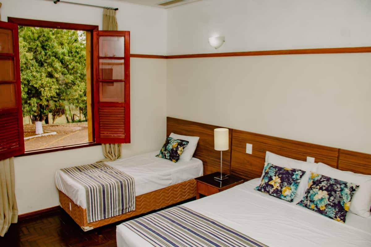 Quarto de hotel com duas camas, janela com venezianas abertas, cabeceira de madeira e mesa de cabeceira com abajur.