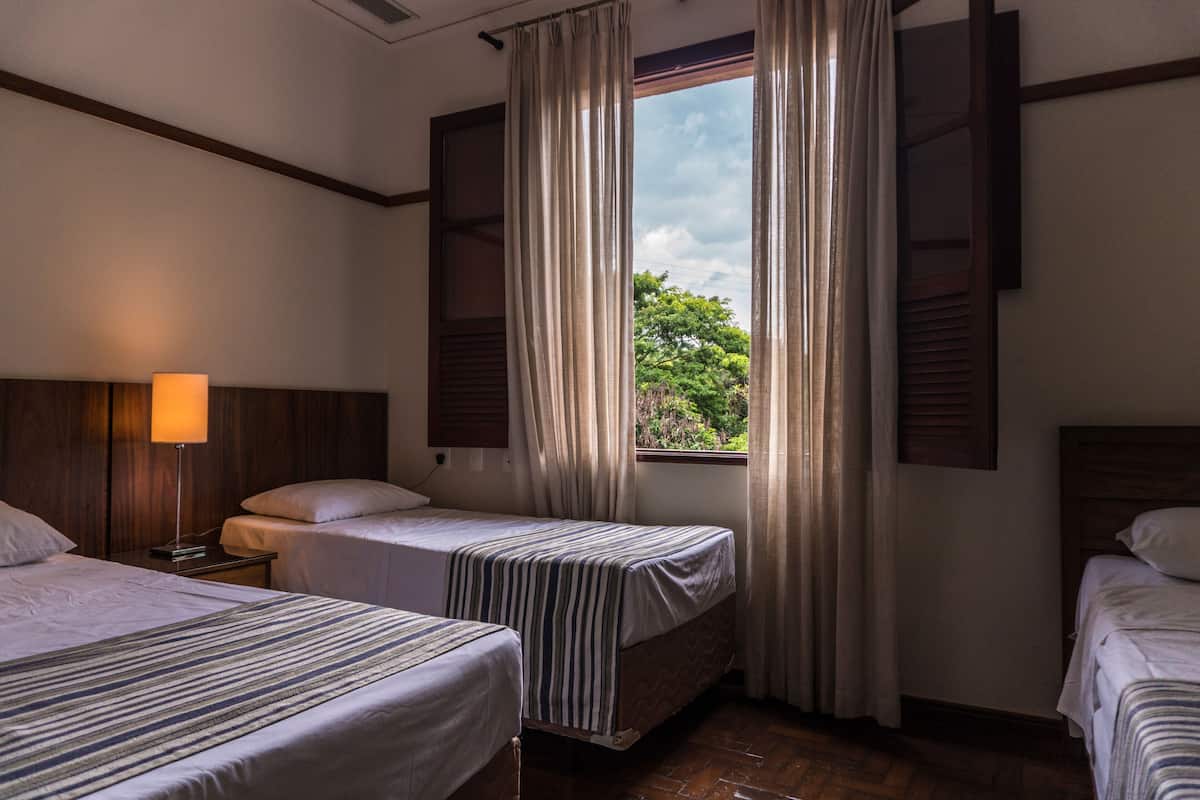 Quarto de hotel com duas camas de solteiro, uma mesa de cabeceira com abajur e uma janela com cortinas.