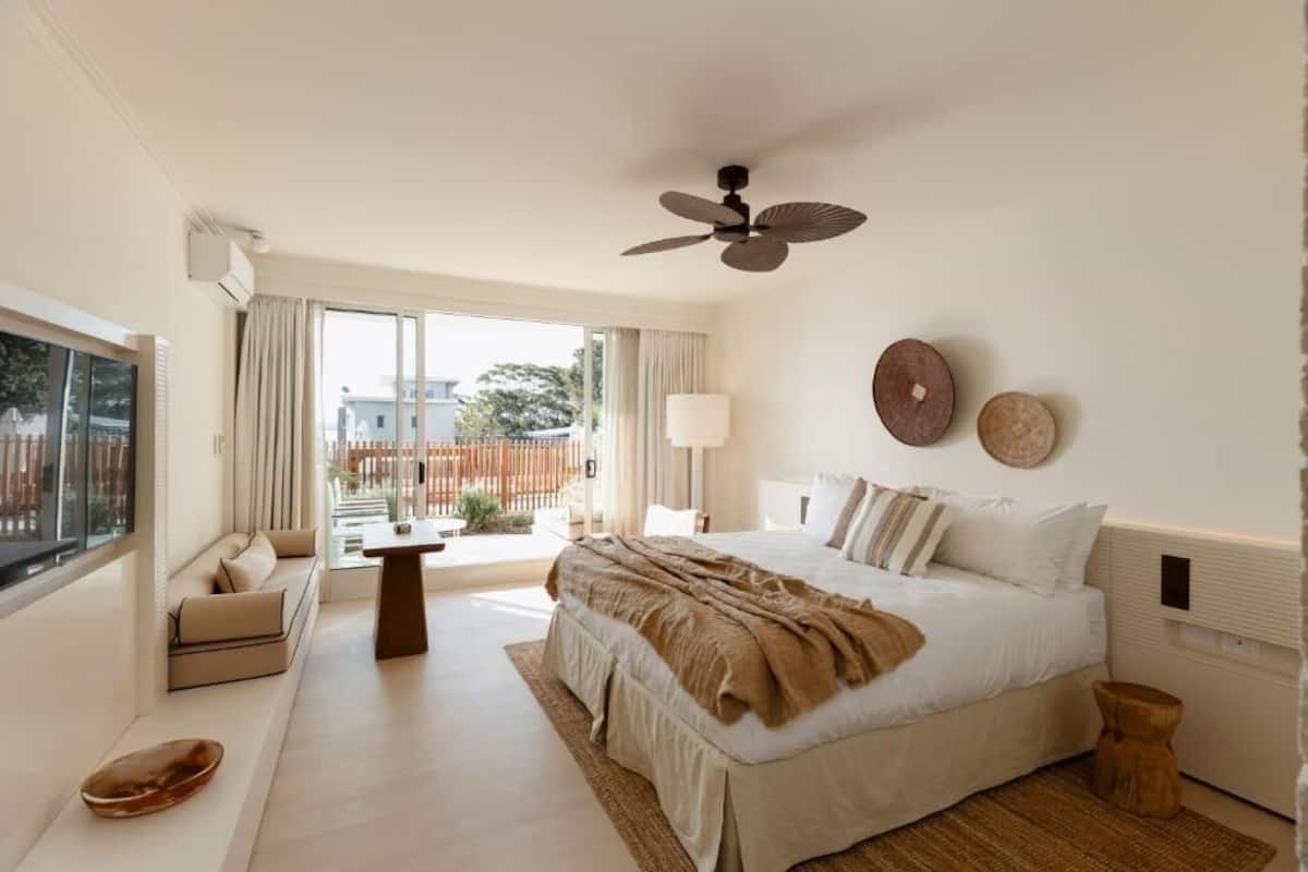 Um quarto espaçoso com uma cama grande, um ventilador de teto, uma janela com cortinas, uma mesa pequena e um espaço para sentar.