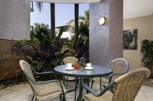 Um pátio com cadeiras de vime e uma mesa, rodeado por plantas e vista para um edifício.