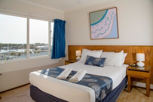 Quarto de hotel com uma cama grande, duas mesas de cabeceira, uma janela com vista para barcos e edifícios, e um quadro na parede.