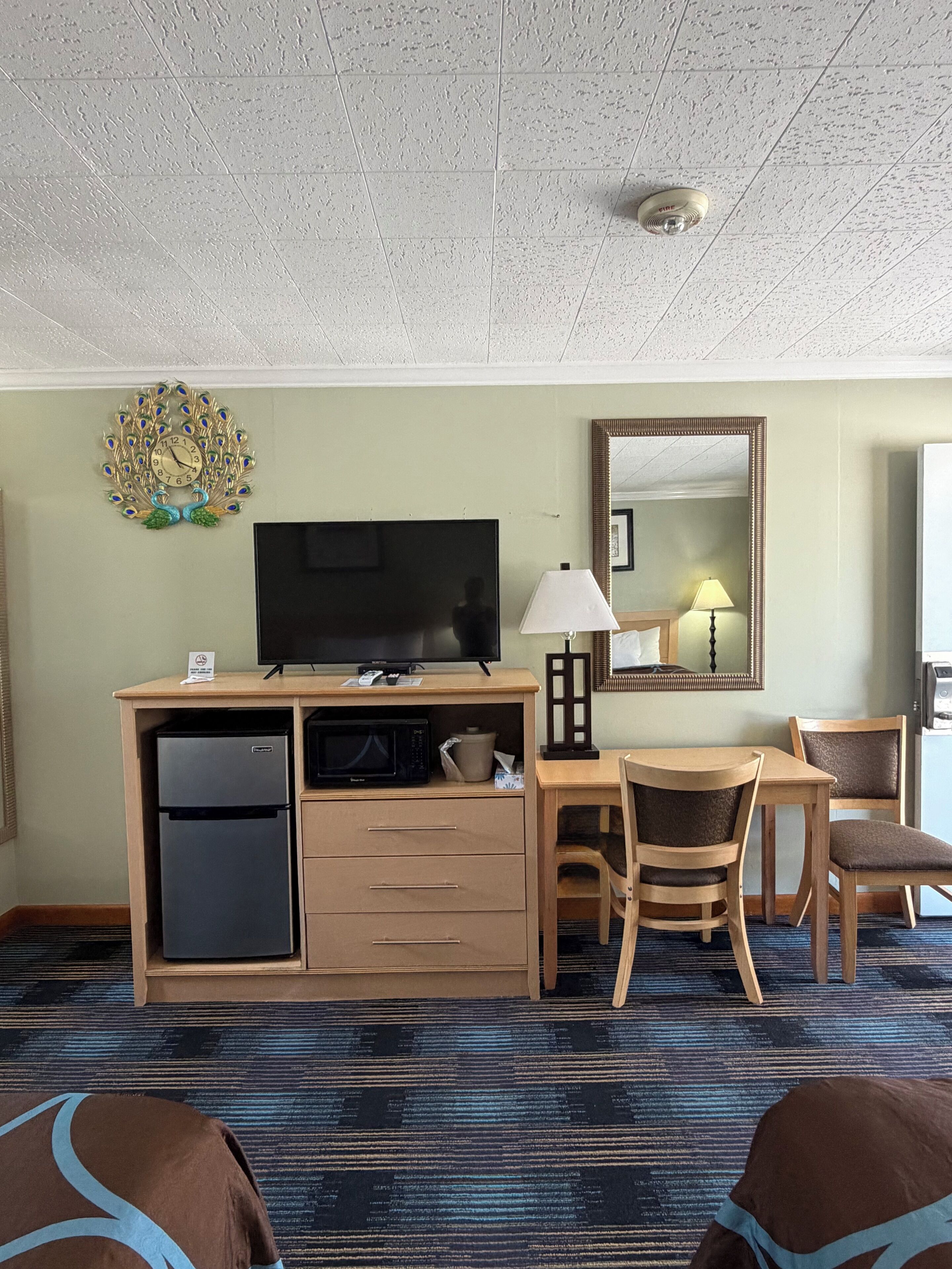 Quarto de hotel com televisão de ecrã plano, micro-ondas, frigorífico, secretária com candeeiro, duas cadeiras e um painel decorativo com tema de pavão.