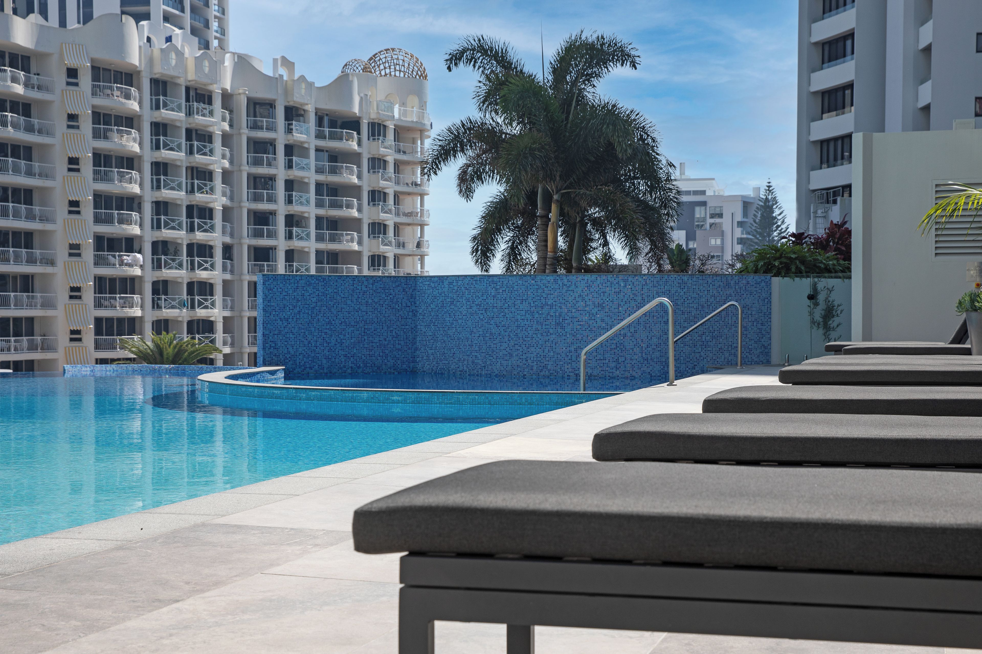 Piscina com espreguiçadeiras, parede revestida de azulejos azuis e um edifício de vários andares com varandas.