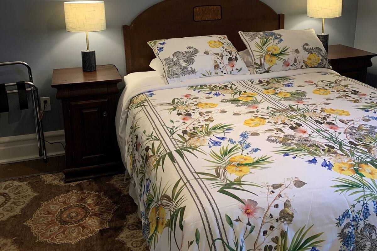 Cama bem arrumada com roupa de cama florida, duas luminárias de cabeceira, um quadro na parede e um tapete estampado.