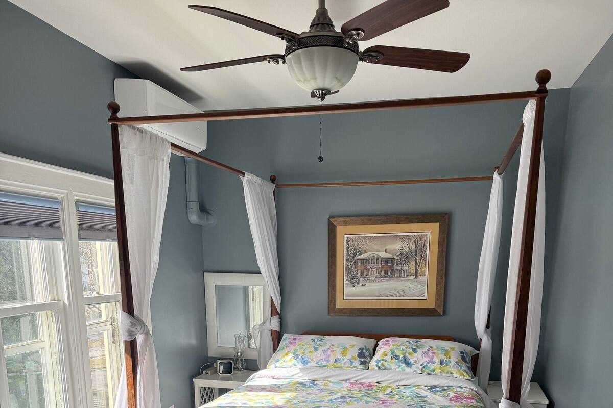Uma cama com dossel e cortinas florais, um ventilador de teto e uma janela grande com cortinas.