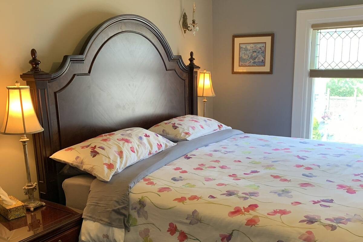 Uma cama bem arrumada com roupa de cama floral, cabeceira de madeira, duas luminárias de mesa e uma mesa de cabeceira com abajur e caixa de lenços.