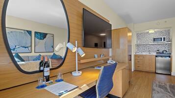 Quarto de hotel moderno com secretária de madeira, espelho grande, televisão de tela plana, área de cozinha com micro-ondas e mesa de jantar.
