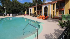 Uma área de piscina com piscina azul cristalina, cadeiras de lounge laranja e um edifício com fachada amarela e varandas.