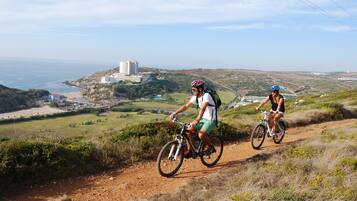 Dois ciclistas em uma trilha de terra com vista para o litoral, incluindo edifícios e vegetação.