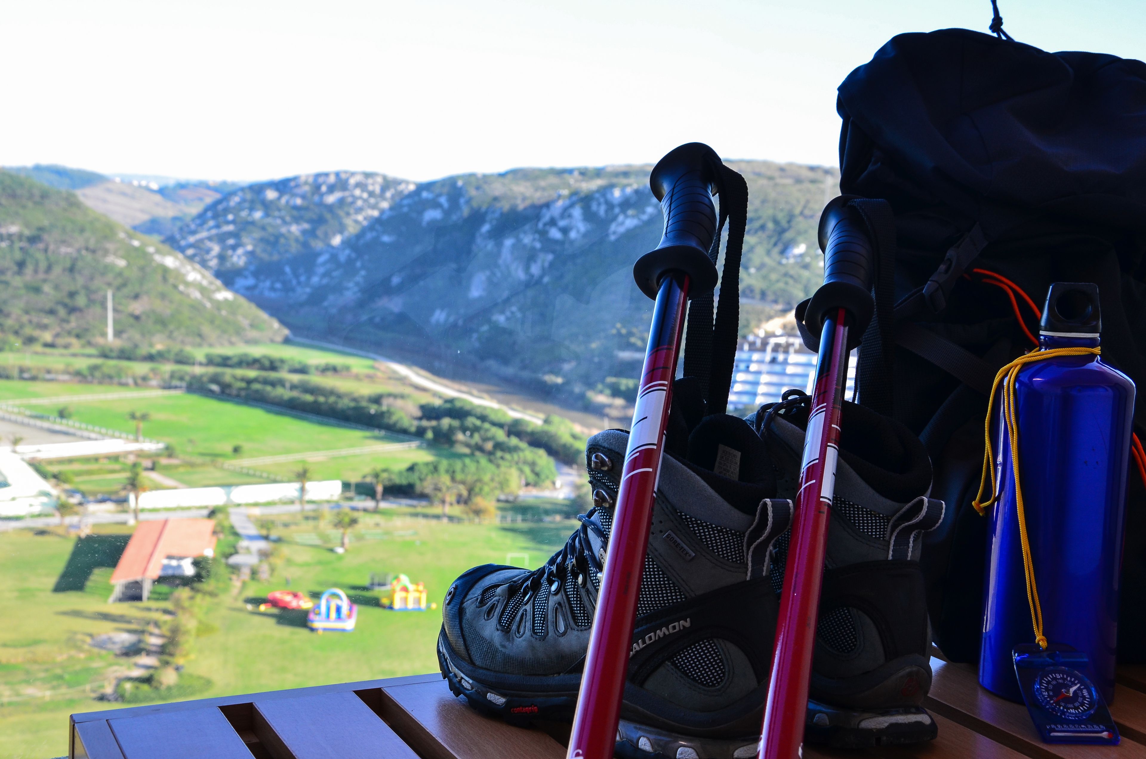 Um par de botas de caminhada, uma bengala e uma garrafa de água azul sobre uma mesa, com vista panorâmica de um vale e montanhas ao fundo.