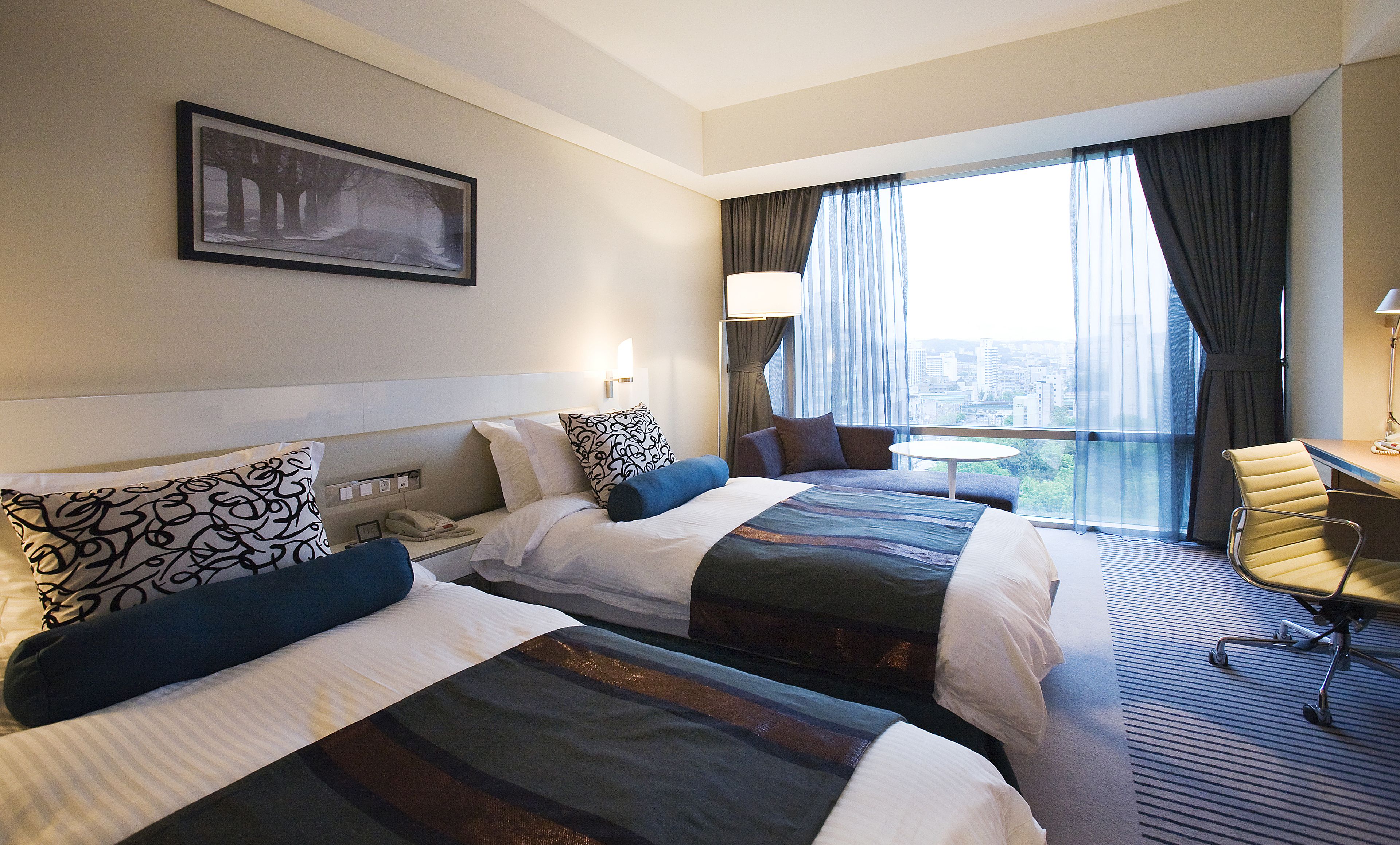 Quarto de hotel com duas camas, uma escrivaninha, uma cadeira, janela com cortinas e vista para a cidade.