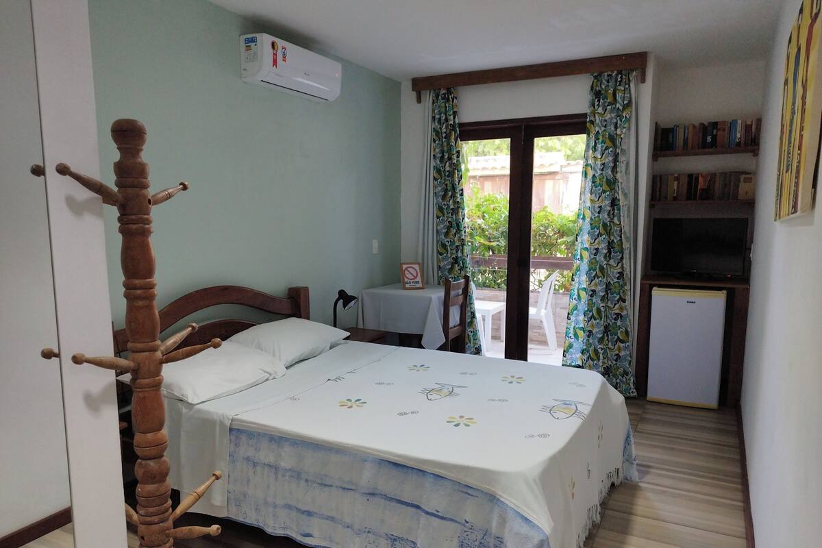 Quarto com cama, porta de madeira, mesa pequena, cadeira, televisão e frigorífico.