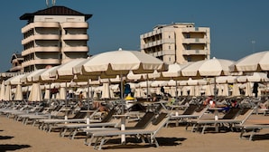 Uma praia com fileiras de espreguiçadeiras sob guarda-sóis, um prédio com várias varandas e uma estrutura em forma de torre ao fundo.