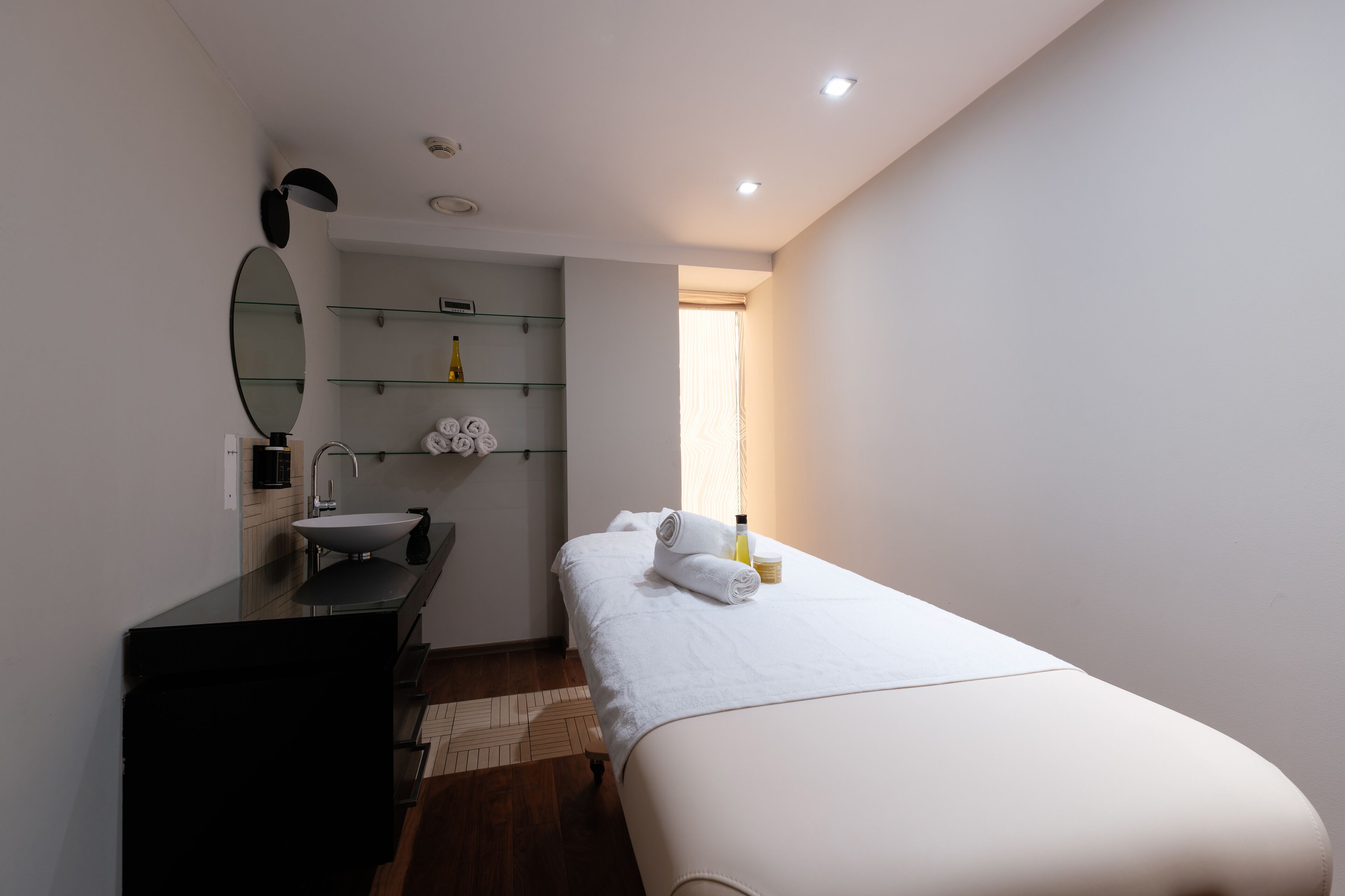 Quarto de hotel moderno com cama, mesa de cabeceira, espelho e banheiro pequeno com pia e porta-toalhas.