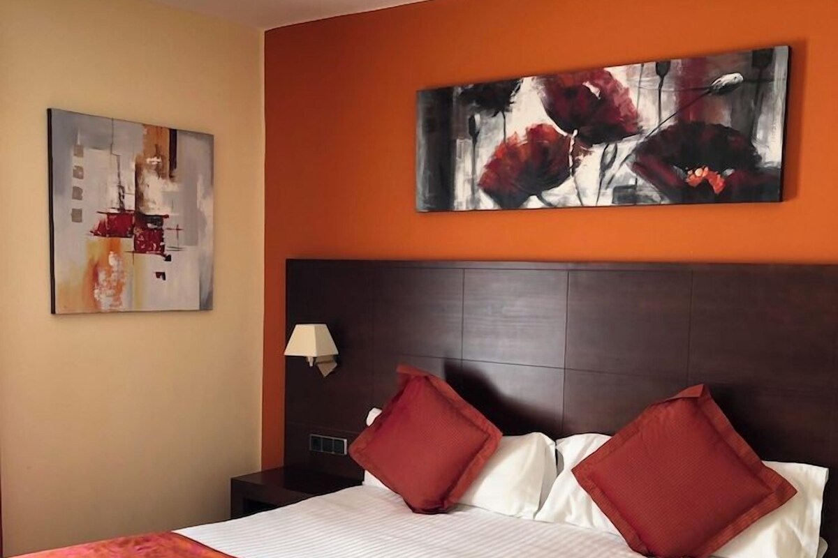 Quarto de hotel com uma cama grande, duas almofadas, uma mesinha de cabeceira, uma luminária e duas obras de arte na parede.