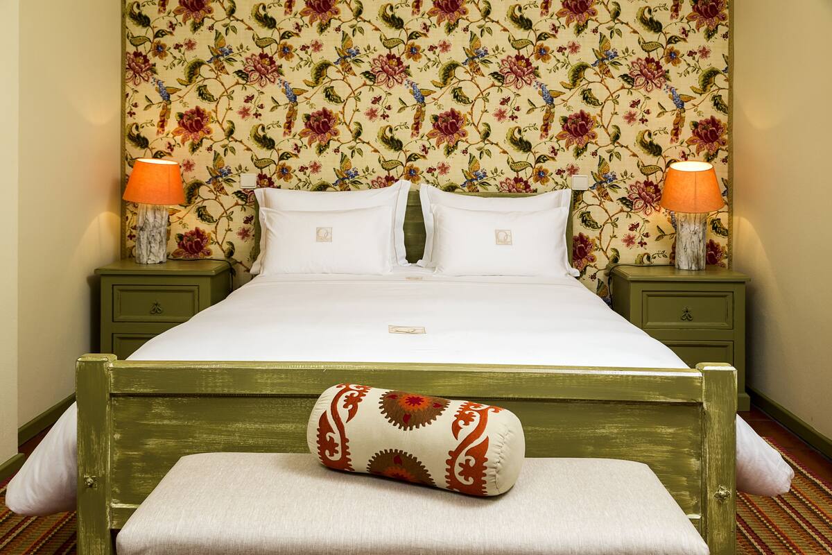 Cama bem arrumada com lençóis brancos, duas luminárias de cabeceira e cabeceira revestida com papel de parede floral.