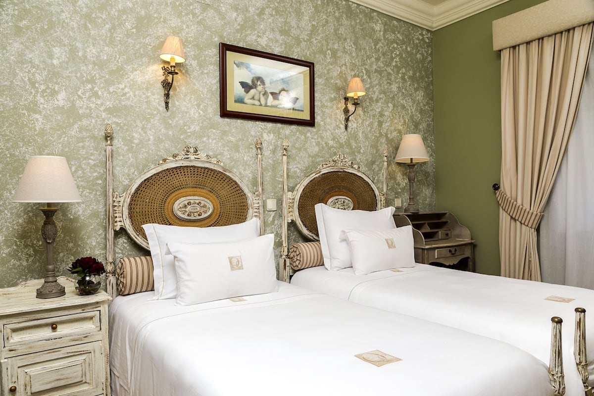 Quarto de hotel com duas camas, cabeceiras elaboradas, arandelas, um quadro emoldurado e uma mesa de cabeceira com abajur.