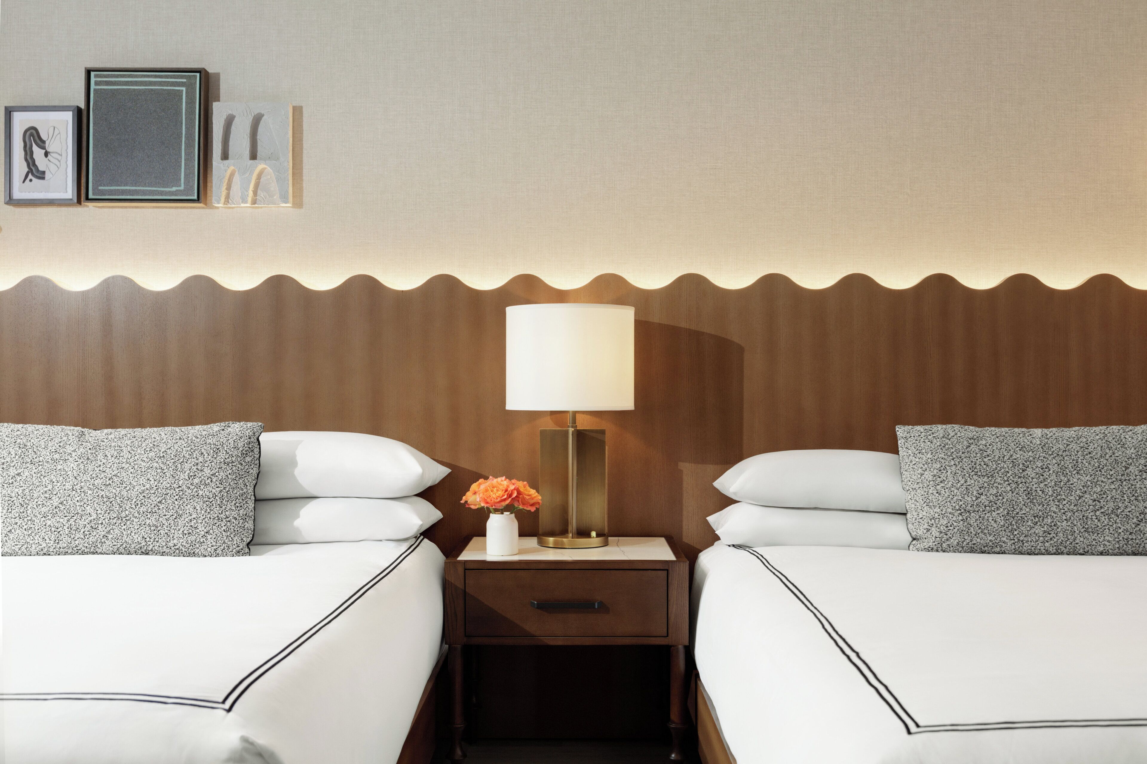 Quarto de hotel com duas camas, mesa de cabeceira com abajur, um vaso com flores e quadros nas paredes.
