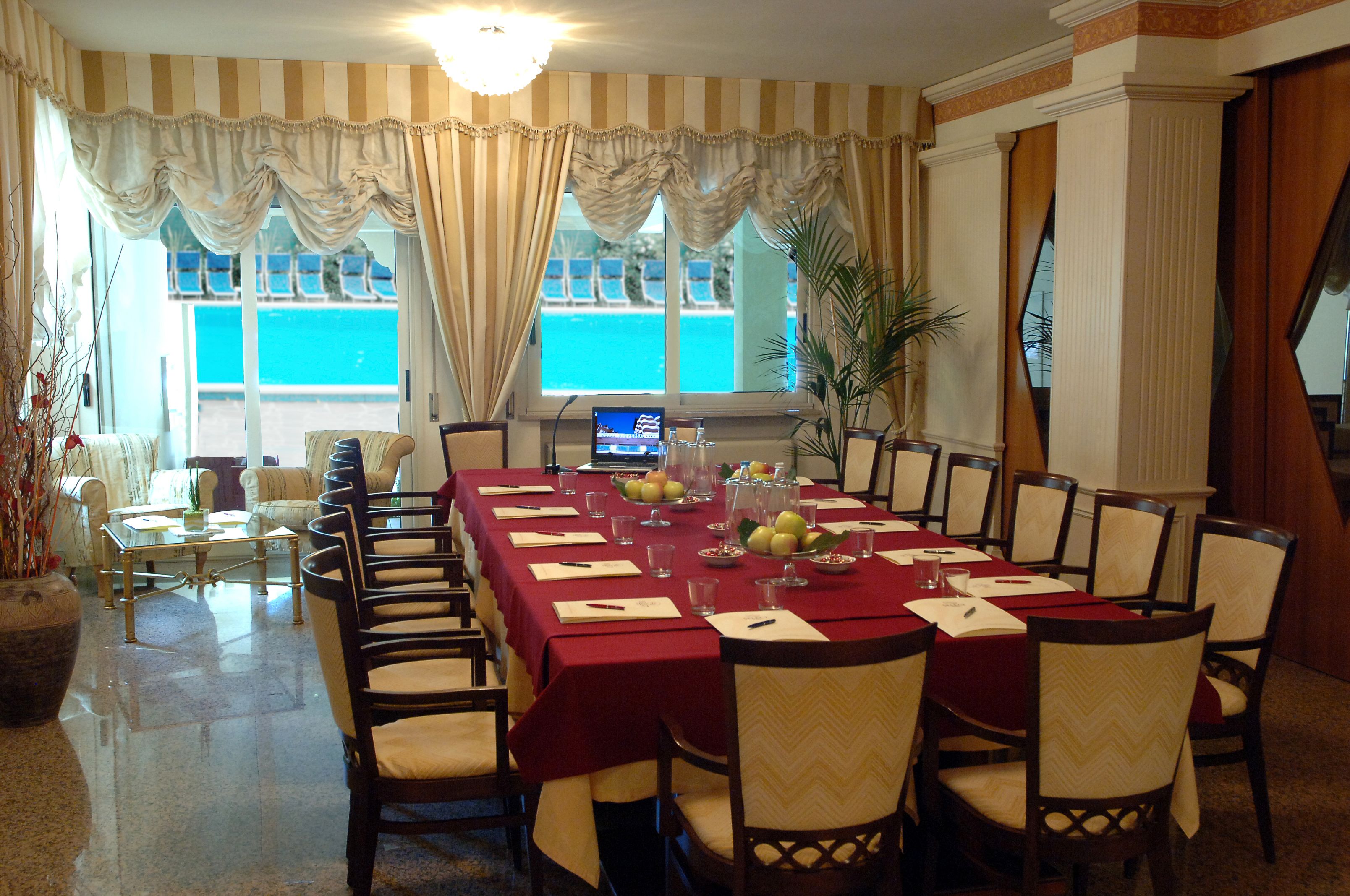 Uma sala de jantar com uma mesa comprida posta para refeição, cadeiras, uma televisão e vista para uma piscina através de grandes janelas.