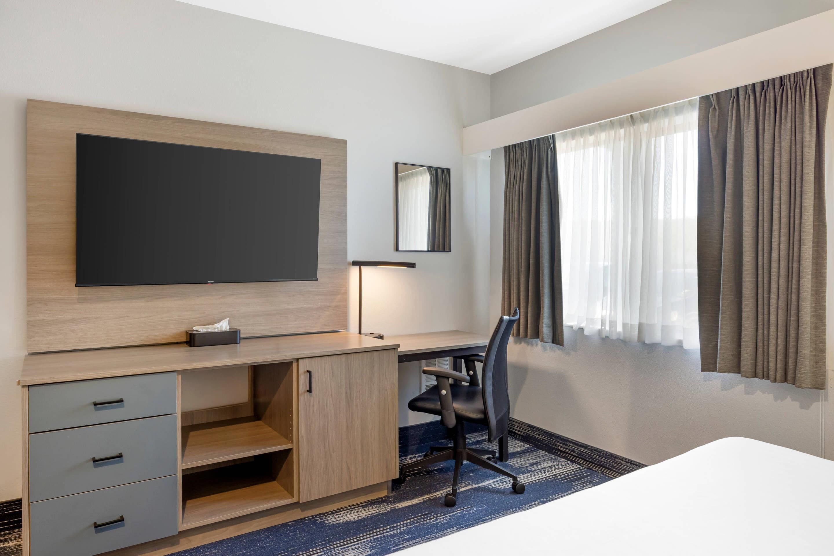 Quarto de hotel com uma cama, uma escrivaninha com cadeira, televisão, espelho e janela com cortinas.