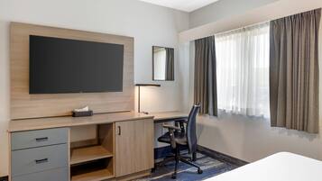 Quarto de hotel com uma cama, uma escrivaninha com cadeira, televisão, espelho e janela com cortinas.