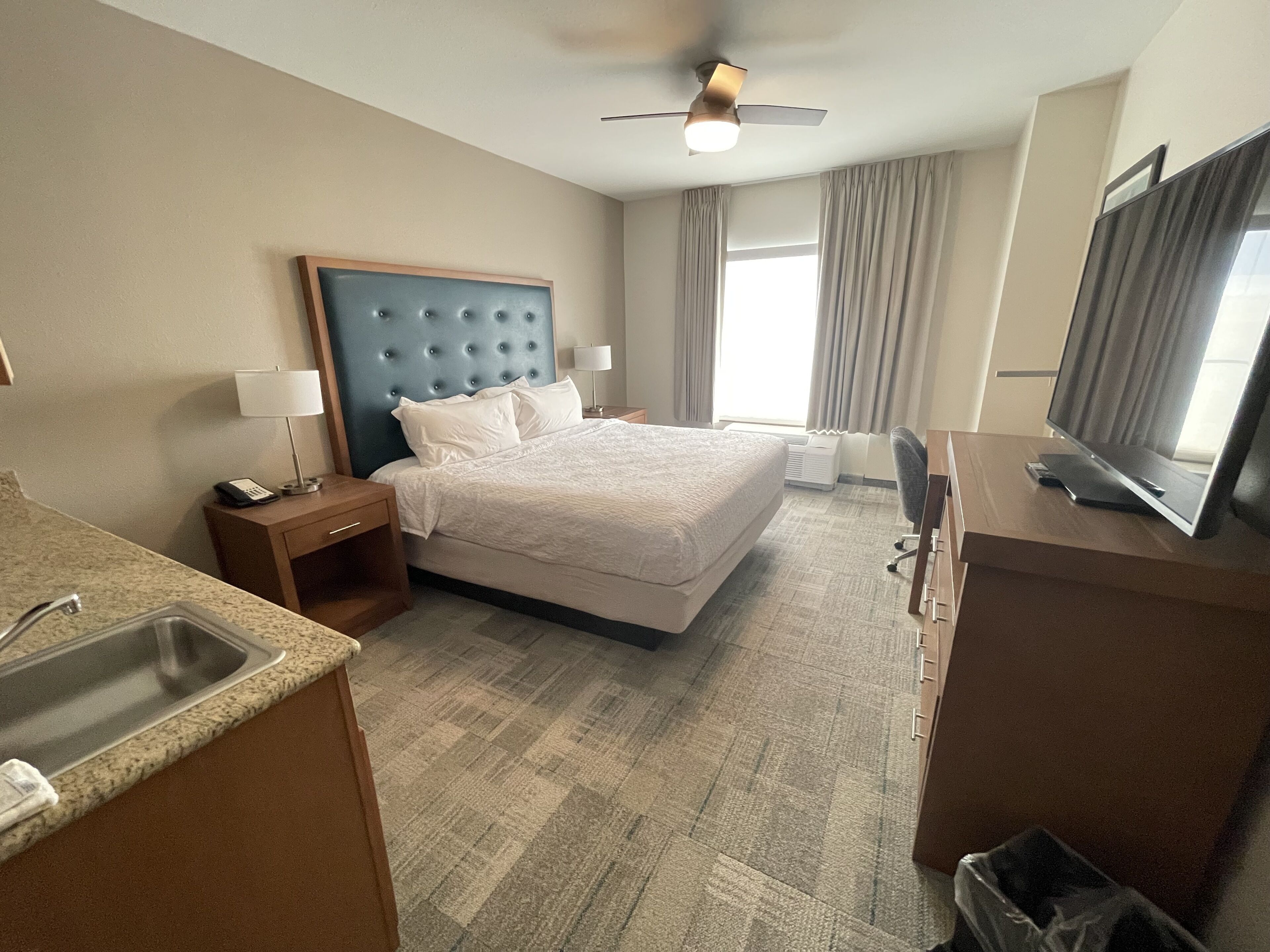 Quarto de hotel com cama, escrivaninha, cadeira, televisão, ventoinha no teto e janela com cortinas.