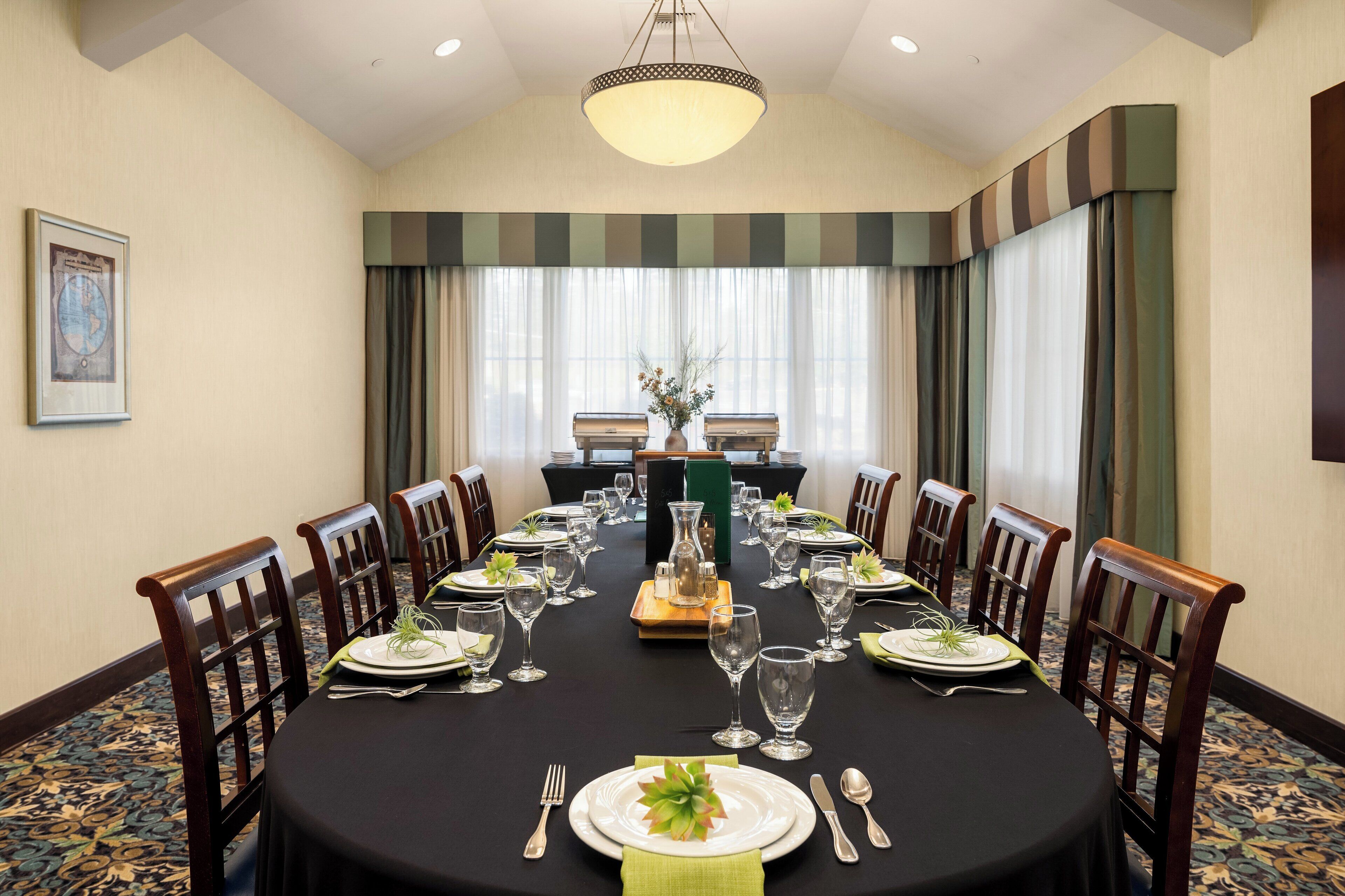 Uma sala de conferências com uma mesa redonda preparada para uma reunião, equipada com cadeiras, um lustre e uma grande janela com cortinas.
