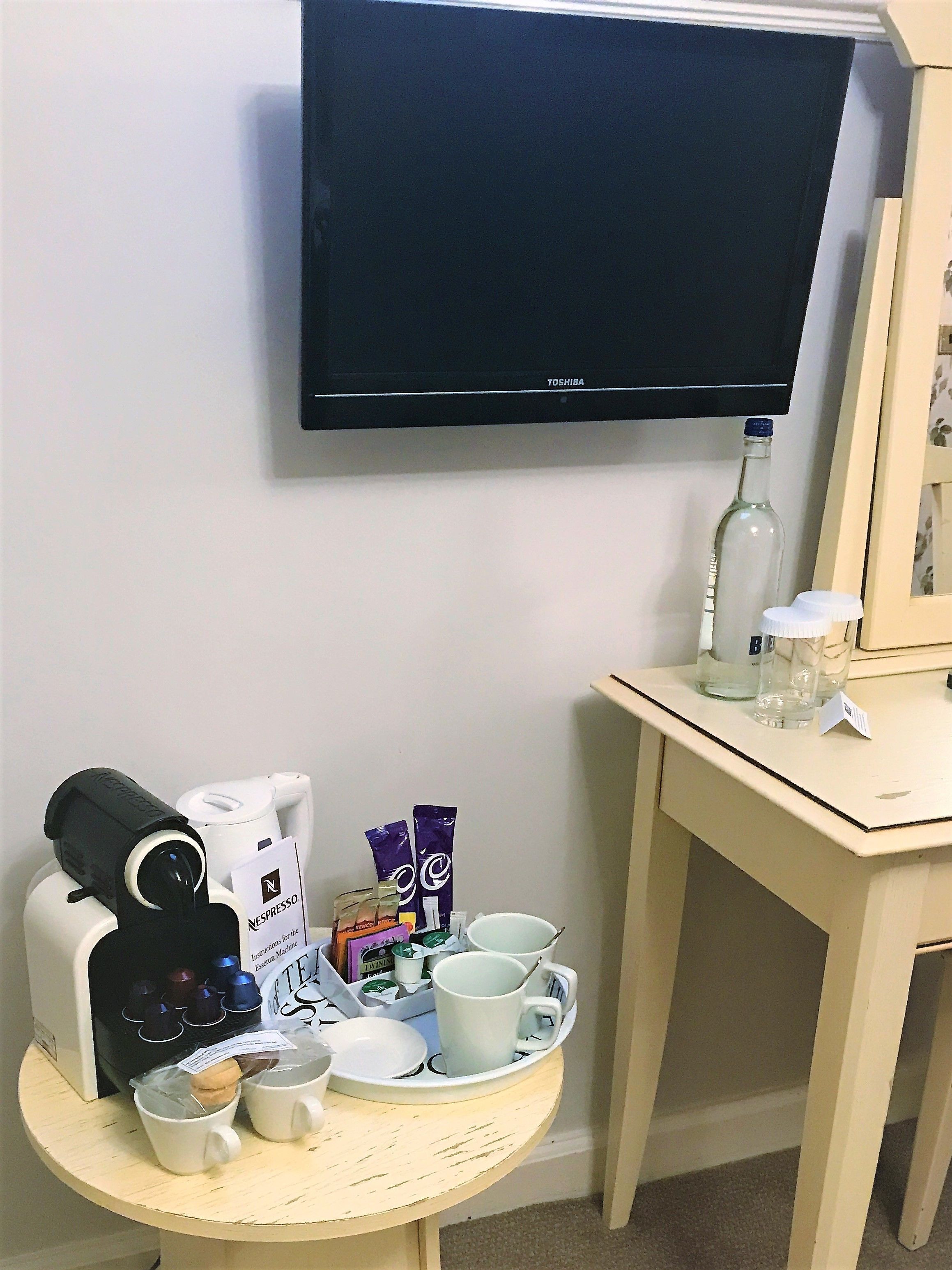 Quarto de hotel com televisão de tela plana na parede, cafeteira, xícaras, uma garrafa e uma pequena mesa com xícara e pires.