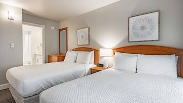Quarto de hotel com duas camas, uma mesa de cabeceira com abajur, uma obra de arte na parede e uma porta para o banheiro.